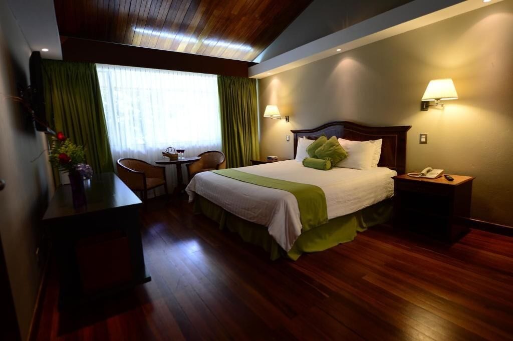 Quarto de hotel com cama, mesa de cabeceira, abajur e escrivaninha com cadeira.