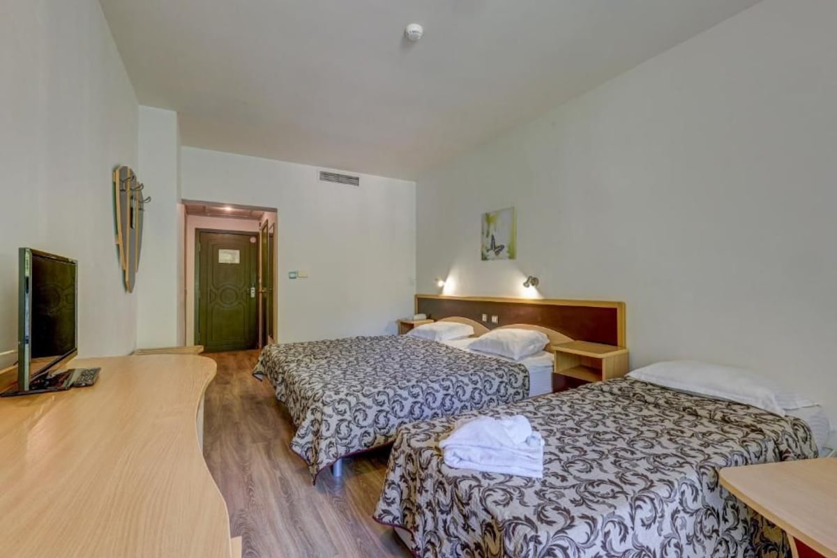 Quarto de hotel com duas camas, uma televisão, uma secretária de madeira e uma porta que dá para outro cômodo.