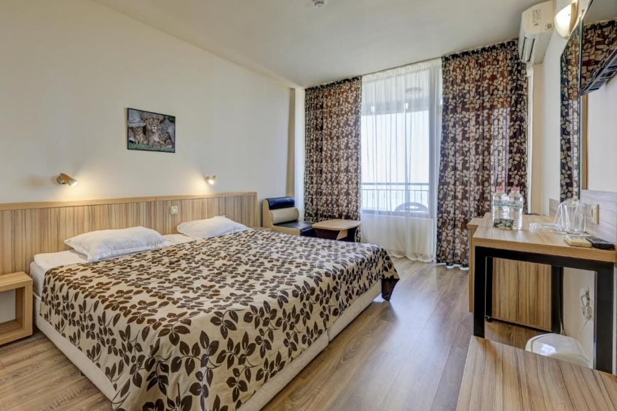Quarto de hotel com cama, escrivaninha, cadeira, mesa pequena, quadro na parede e janela com cortinas.