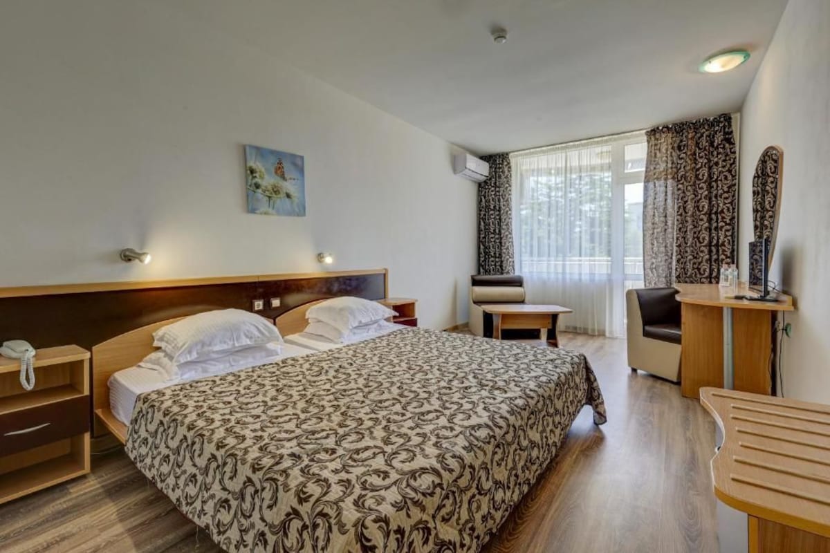 Quarto de hotel com cama, mesa de cabeceira, cadeira, escrivaninha e um quadro na parede.