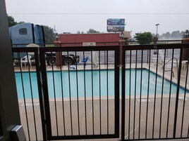 Uma piscina com entrada protegida por grades, um prédio vermelho ao fundo e um outdoor.