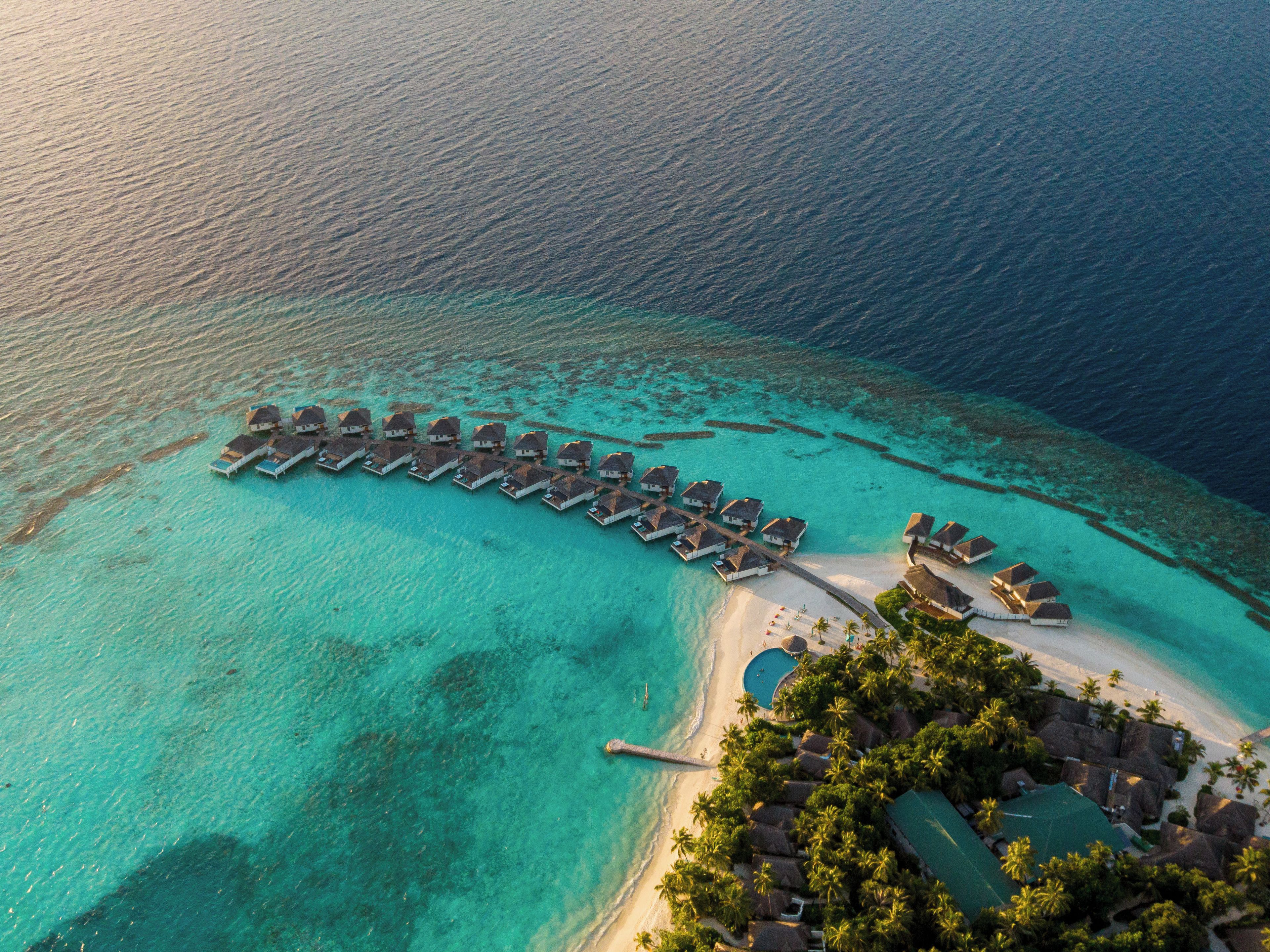 Vista aérea de um resort com bangalôs sobre a água dispostos em semicírculo, uma praia de areia e uma pequena área de piscina.