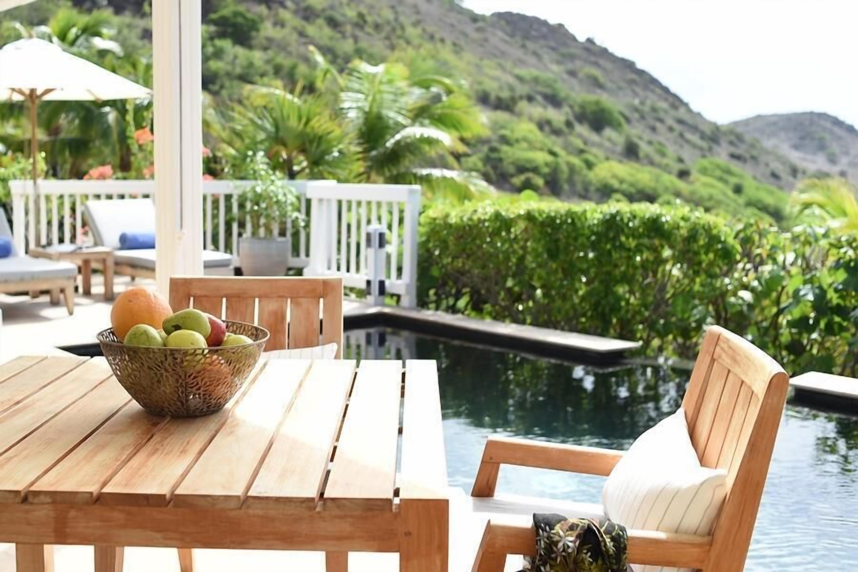 Uma mesa de madeira com uma cesta de frutas, uma cadeira à beira da piscina e vista para uma paisagem verde exuberante.