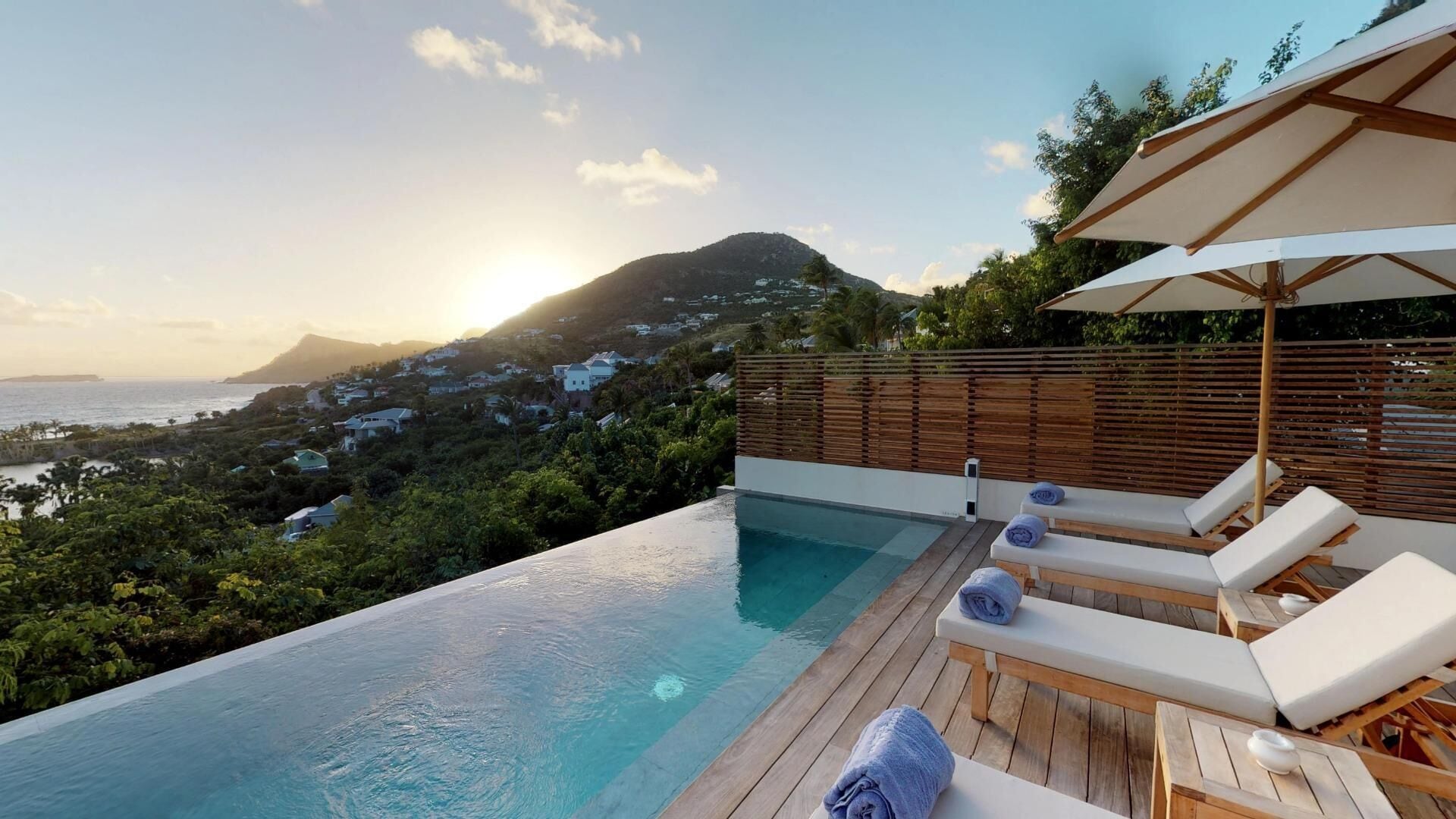 Piscina infinita com deck de madeira, espreguiçadeiras e vista para uma cidade costeira e montanhas.