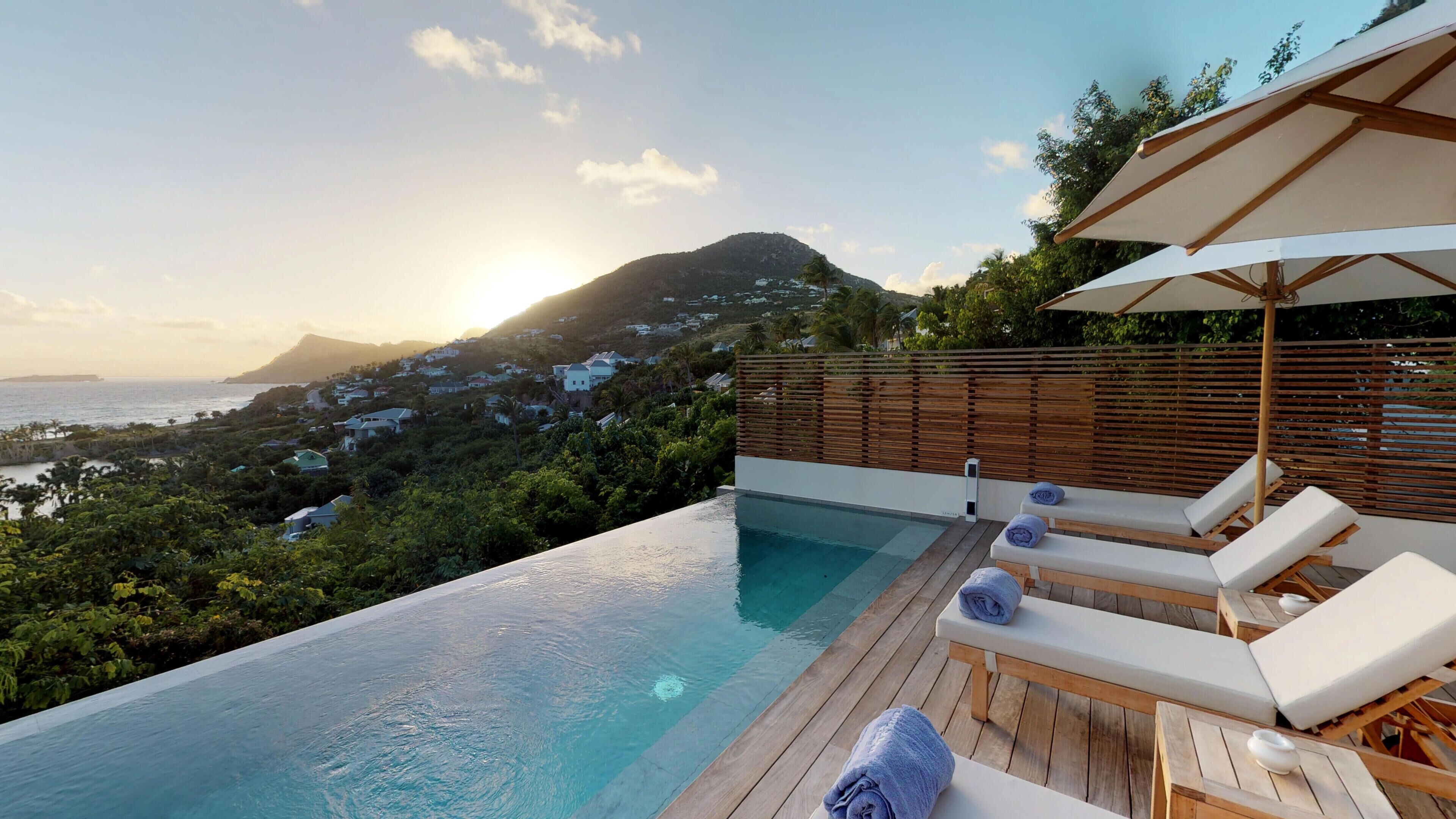 Piscina infinita com vista para o oceano e montanhas, deck de madeira, espreguiçadeiras e cerca de madeira.