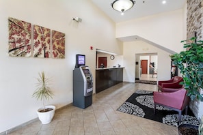 Um hall de entrada com um caixa eletrônico, balcão de recepção, relógio, escada e uma planta em vaso.