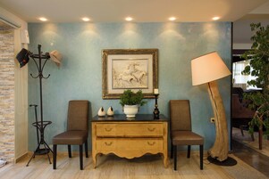 Quarto com cômoda de madeira, duas cadeiras, luminária de chão, porta-roupas e uma obra de arte emoldurada na parede.