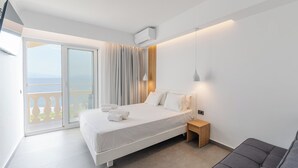 Quarto moderno com cama grande, mesa de cabeceira de madeira, sofá cinza e varanda com cortinas.