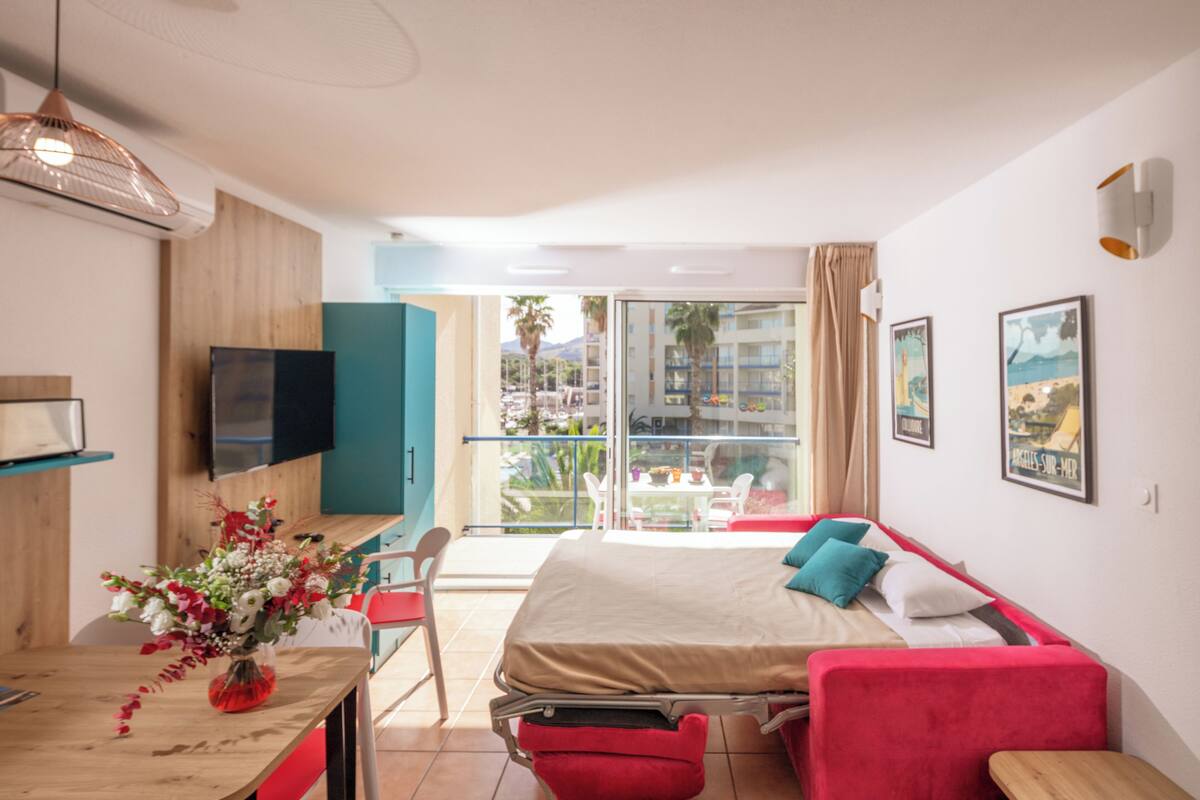 Quarto de hotel com cama, escrivaninha, cadeira, televisão e varanda com vista para palmeiras e edifícios.