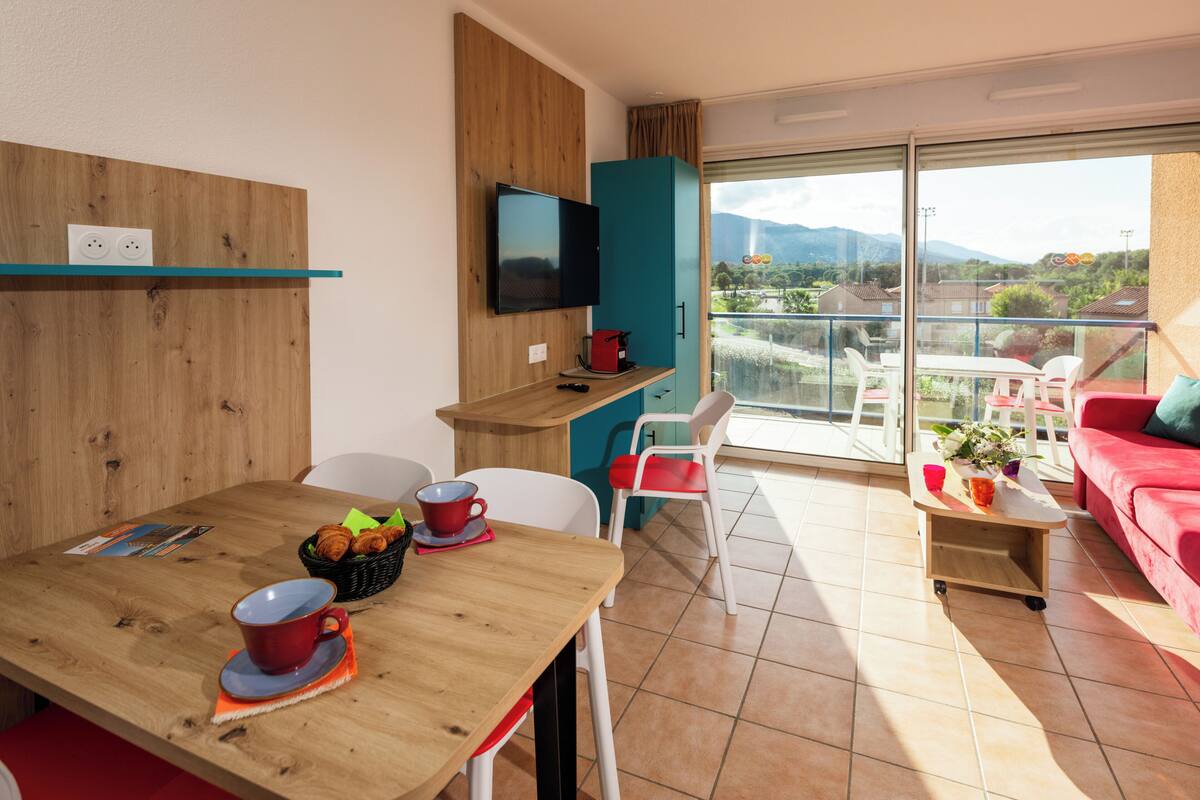 Quarto moderno com mesa de madeira, cadeiras, televisão e varanda com sofá vermelho.