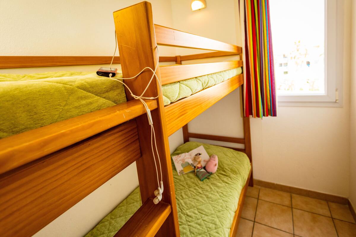 Uma cama beliche com colcha verde, uma mesinha de cabeceira com um livro e um brinquedo, e uma janela com cortinas coloridas.