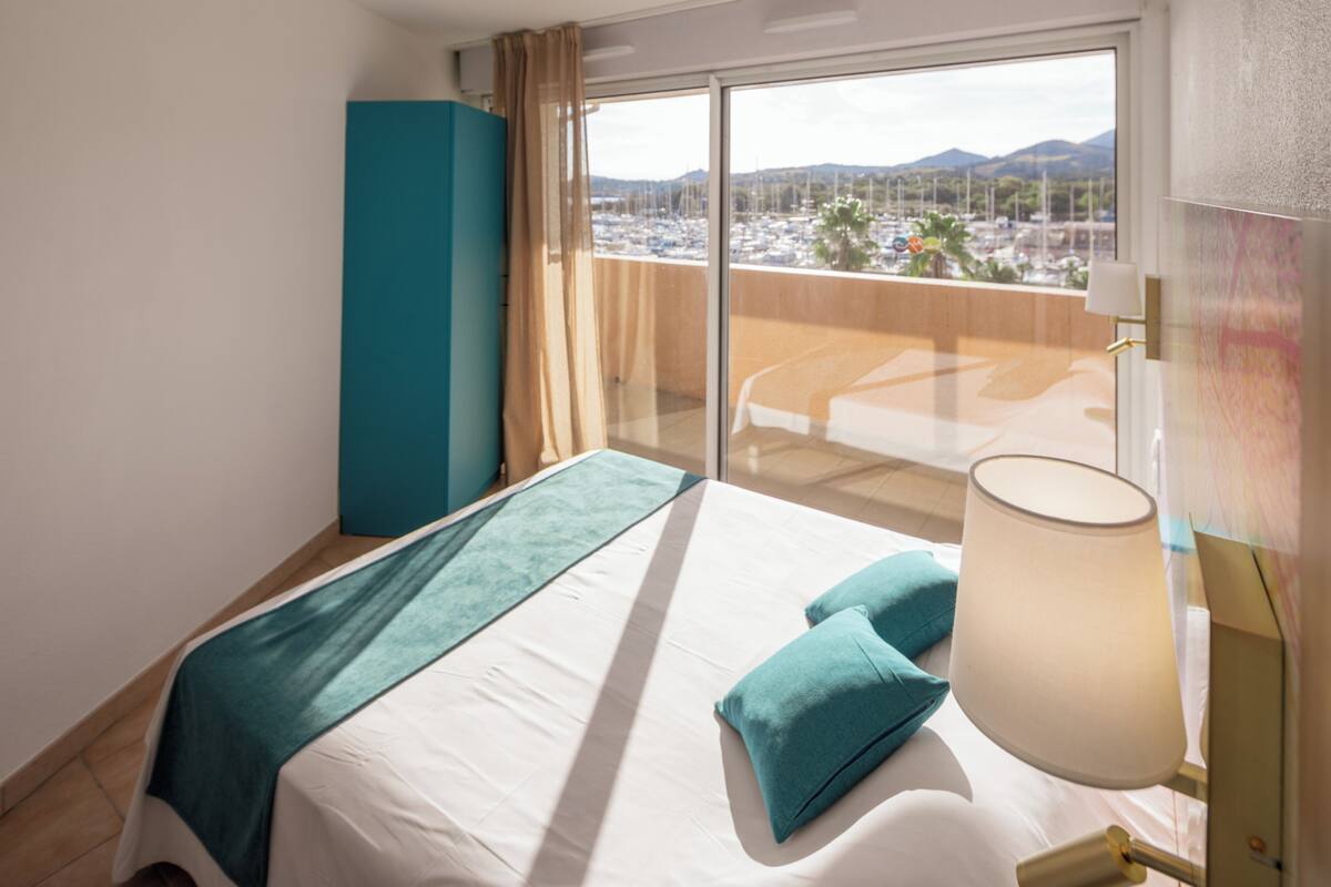 Quarto de hotel com cama, varanda com vista para um estacionamento e montanhas, e uma janela grande com cortinas.