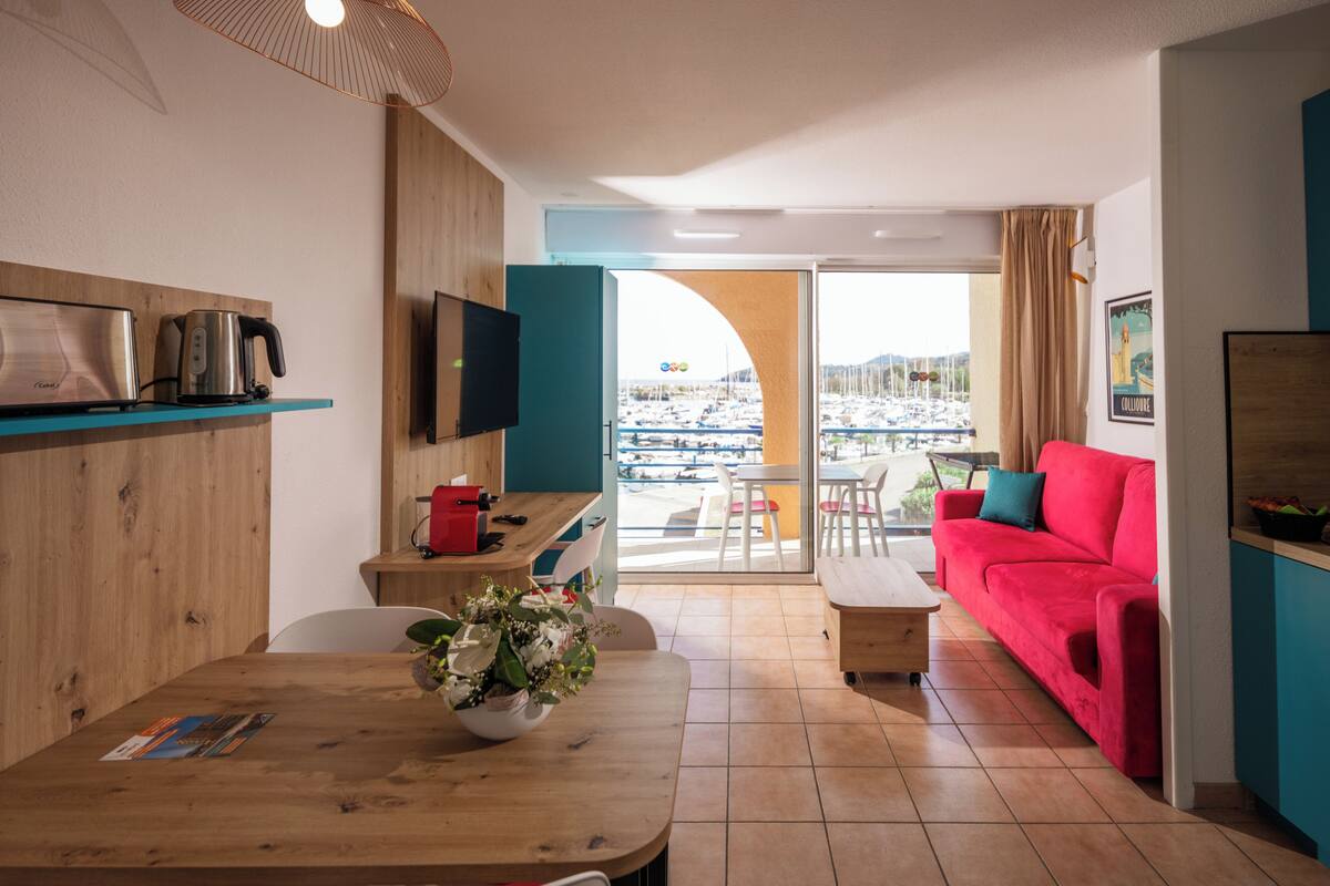 Uma sala de estar moderna com um sofá vermelho, uma mesa de jantar de madeira com um vaso de flores, uma área de cozinha com uma chaleira e uma TV na parede.