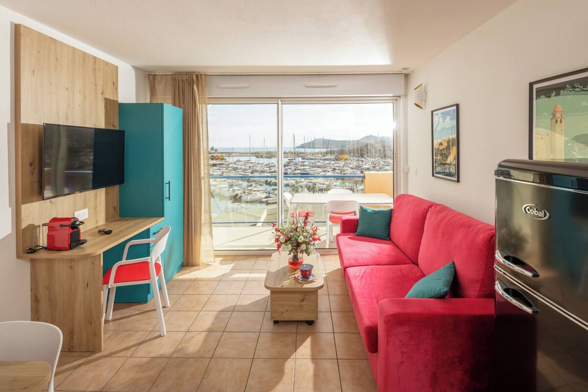 Uma sala de estar moderna com um sofá vermelho, uma TV de tela plana, uma geladeira Cobalt, uma escrivaninha de madeira e vista para um porto através de grandes portas de vidro.