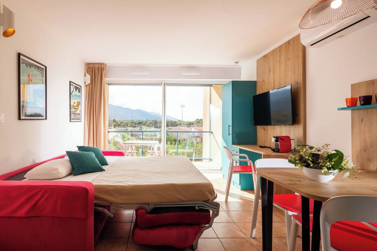 Quarto de hotel com cama, escrivaninha, televisão e varanda com vista.
