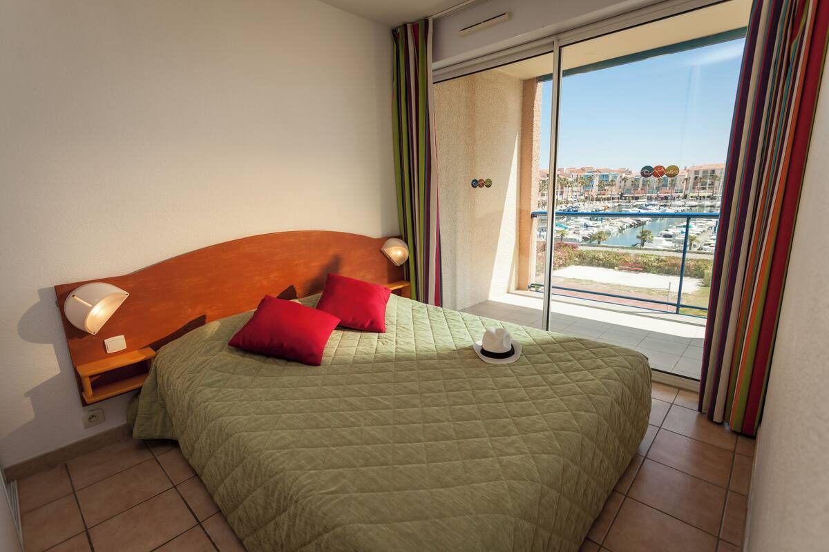 Quarto com uma cama grande, mesa de cabeceira, luminária e vista para um porto desportivo através de uma porta de correr de vidro.