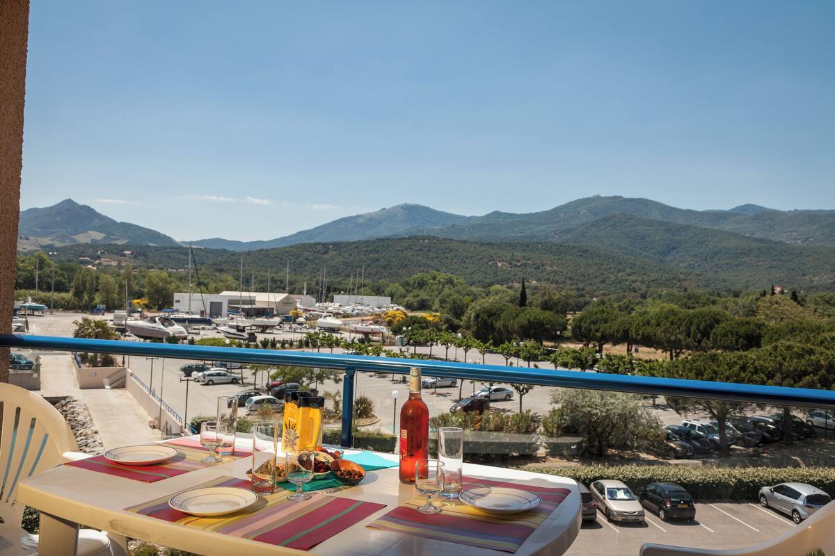 Mesa de varanda posta com pratos, copos e uma garrafa, com vista para um estacionamento e montanhas.
