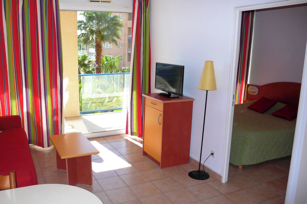 Quarto de hotel com um sofá vermelho, uma mesinha, uma TV sobre um móvel de madeira, um abajur e uma cama com almofadas vermelhas.