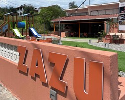 Um parquinho com escorregadores e estruturas de escalada, um edifício com área de estar externa e uma placa com a inscrição 'LAZU'.