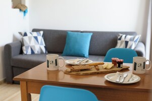 Uma área de refeições com uma mesa de madeira, pratos, talheres e uma caneca. Um sofá cinza com almofadas estampadas está ao fundo.
