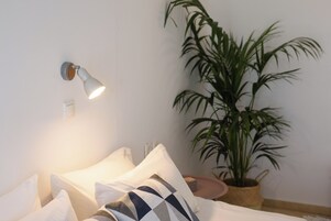 Uma cama com almofadas, uma luminária na parede e um vaso com planta.
