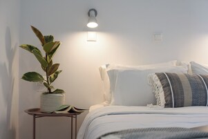 Quarto com uma cama, uma mesinha de cabeceira com uma planta e um candeeiro fixado na parede.