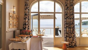 Quarto com vista para o mar, mesa de buffet com diversos alimentos e uma pequena mesa com uma única cadeira.