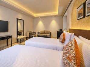 Quarto de hotel com duas camas, uma escrivaninha, uma cadeira, um sofá, uma televisão e um espelho.