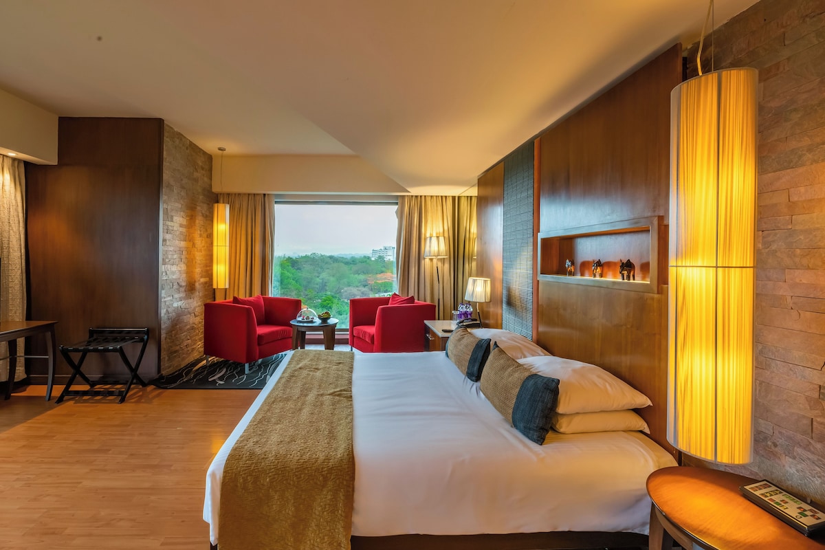Quarto de hotel com uma cama grande, duas poltronas vermelhas, uma mesinha e vista para o exterior.