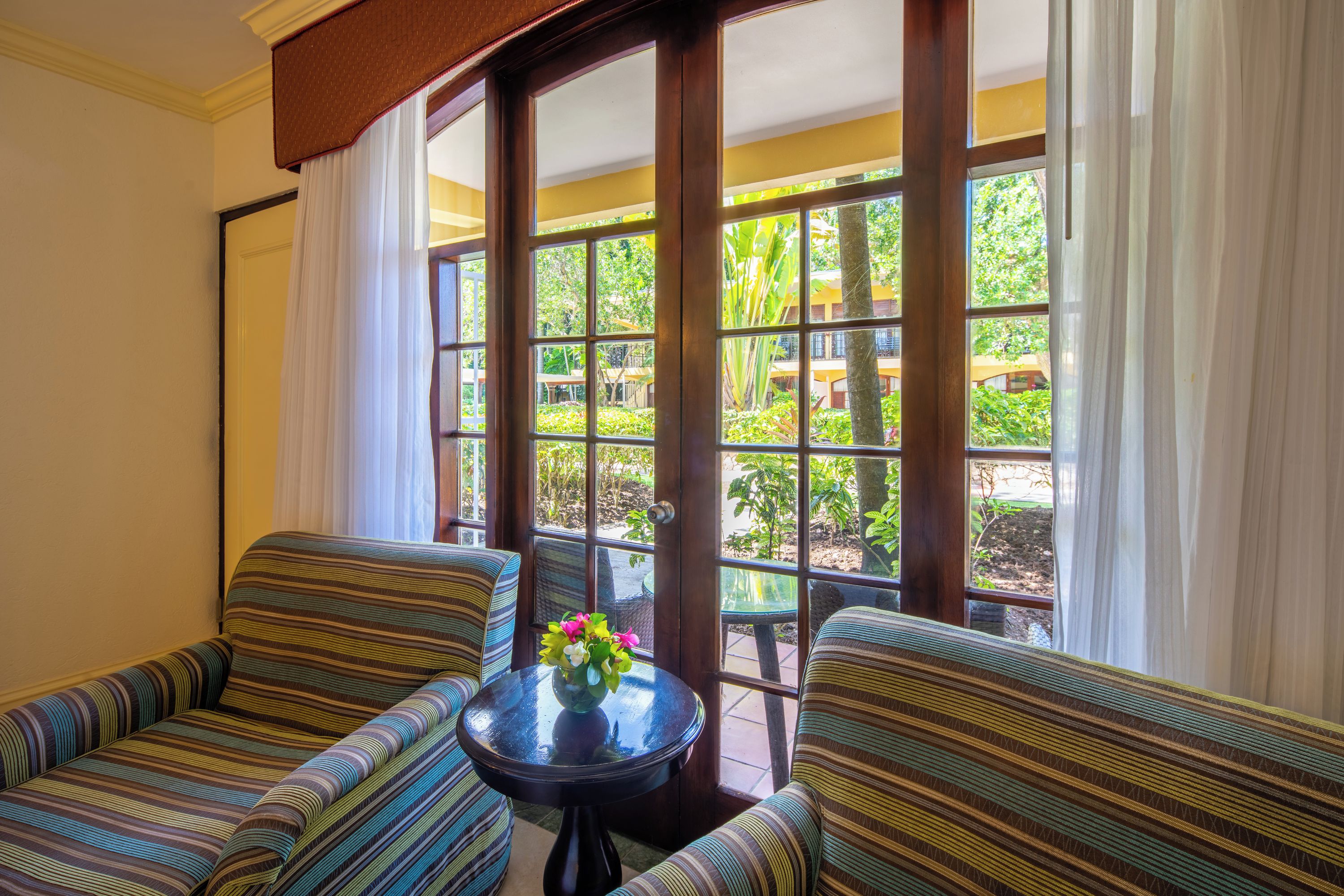 Quarto com dois sofás listrados, uma mesinha redonda com um vaso de flores e uma janela grande com vista para o verde.