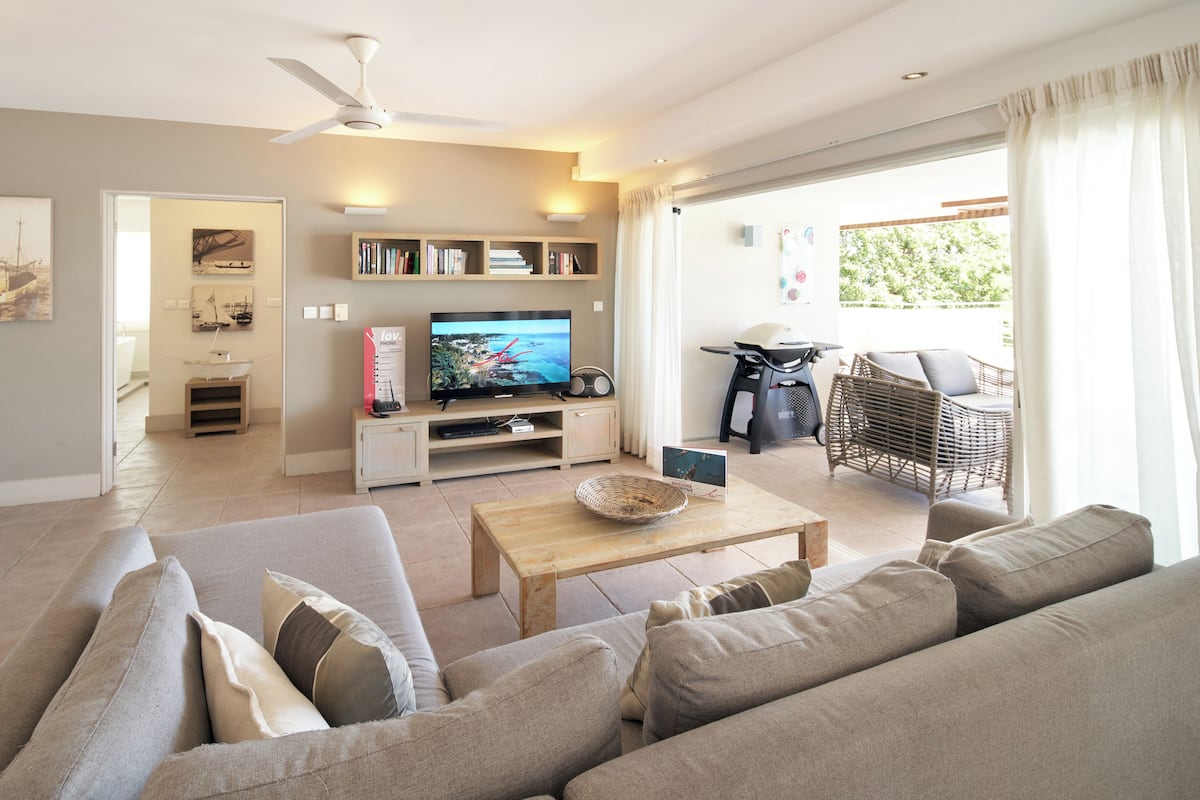 Uma sala de estar com televisão, sofá, mesa de centro e varanda com mobiliário de vime.