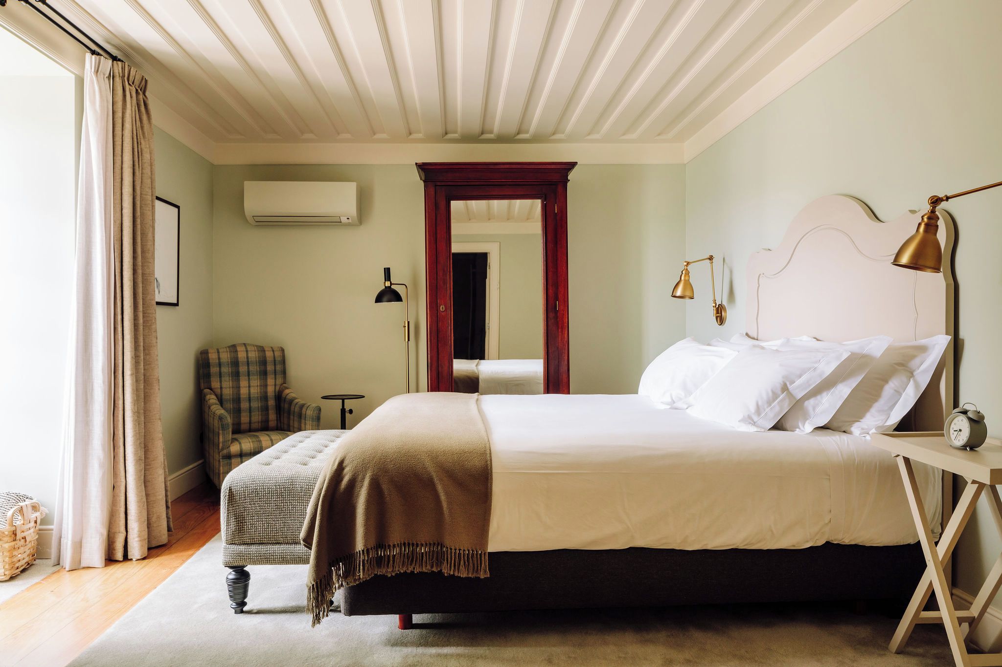 Quarto bem arrumado com cama, poltrona, mesa de cabeceira com luminária e ar condicionado fixado na parede.