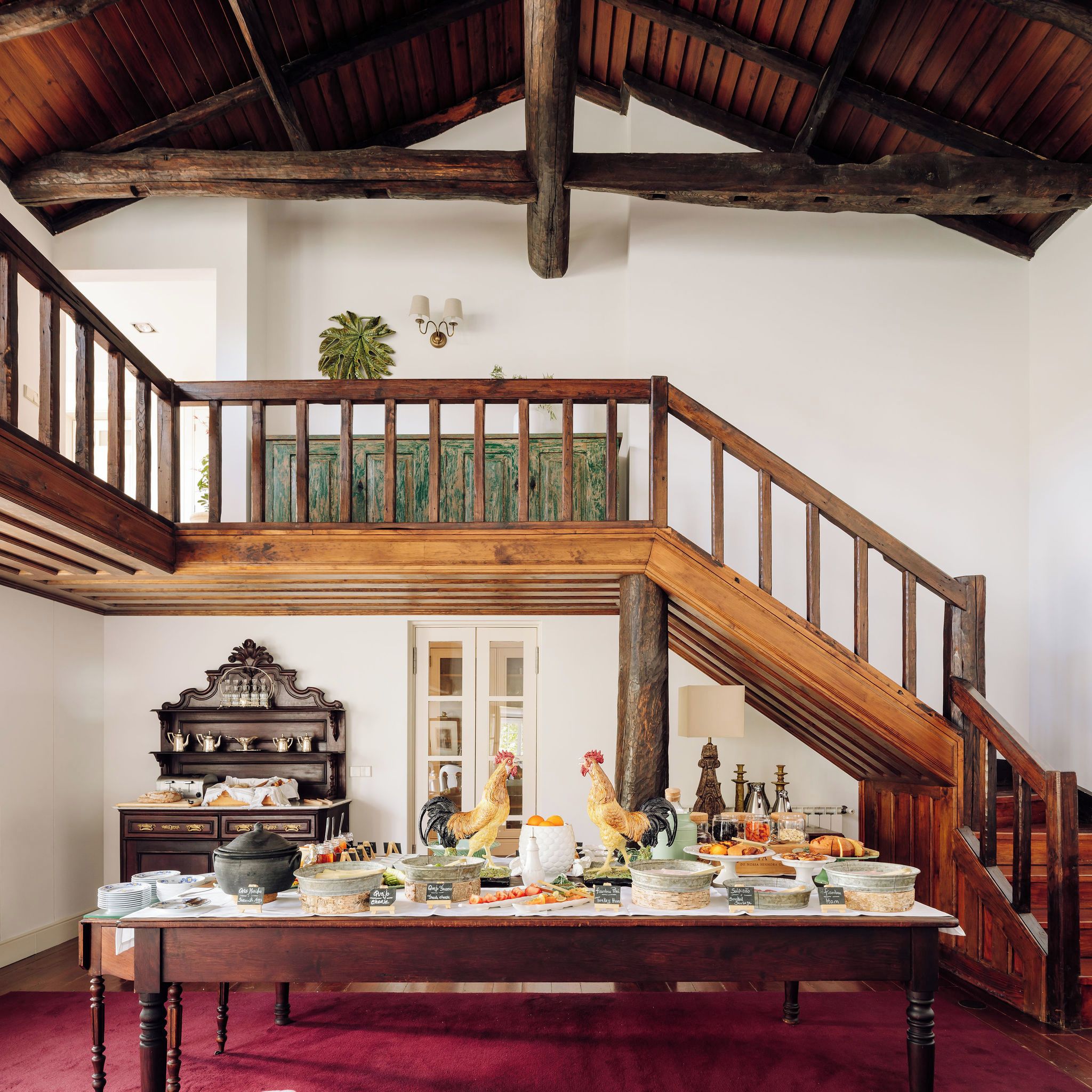 Uma área de refeições com uma escada de madeira, mesa de buffet com alimentos e uma estátua decorativa de galo.
