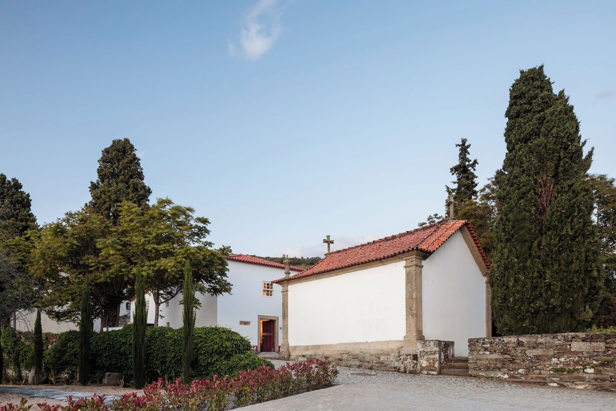Um edifício branco com telhado de telhas vermelhas, rodeado por árvores e um muro de pedra.
