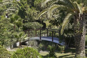 Uma ponte curva sobre um jardim com diversas plantas e uma palmeira.