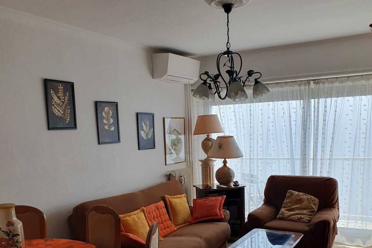 Uma sala de estar com um sofá marrom, uma mesa de centro, uma mesa de jantar com toalha de mesa florida e um abajur.
