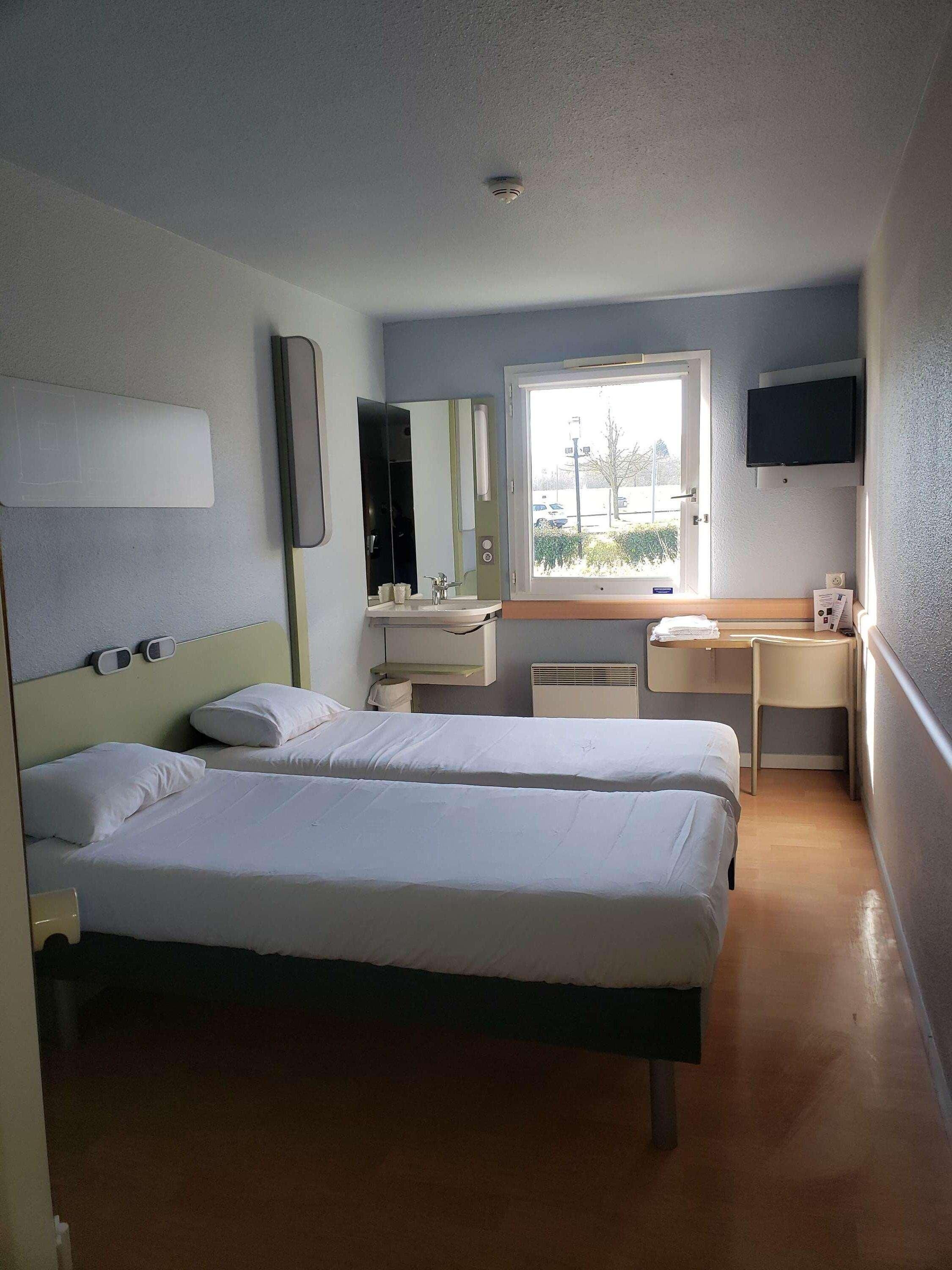 Quarto de hotel com duas camas, uma escrivaninha, televisão, janela e banheiro.