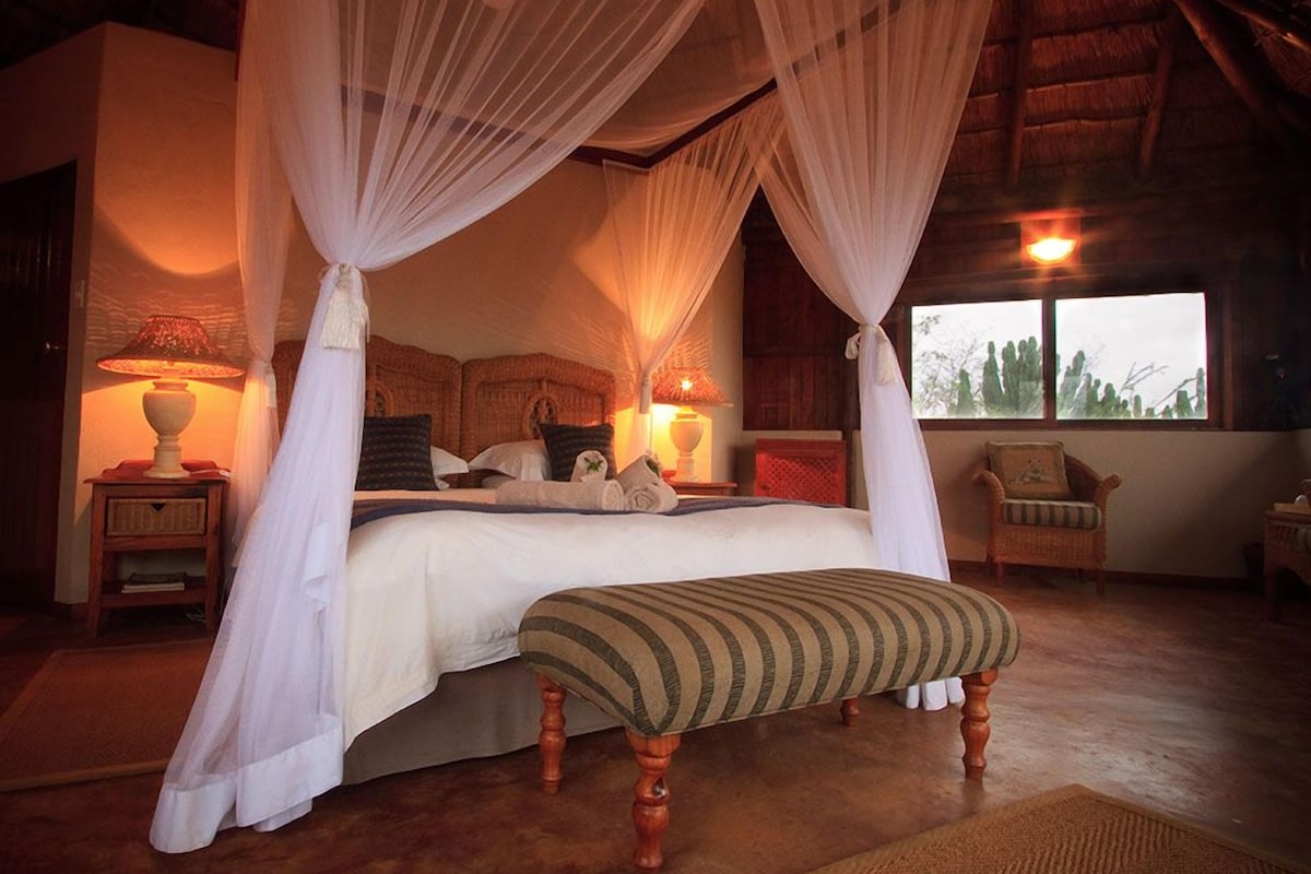 Quarto com cama de dossel, luminárias de cabeceira, um banco e vista para árvores pela janela.