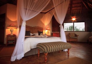 Quarto com cama de dossel, luminárias de cabeceira, um banco e vista para árvores pela janela.