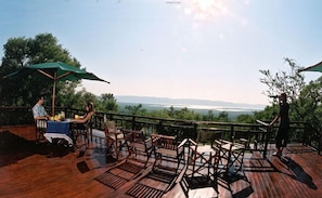 Um deck de madeira com área de estar ao ar livre, uma mesa com bebidas e vista para a água e montanhas ao longe.