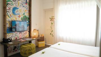 Quarto de hotel com duas camas, uma secretária com telefone e papéis, uma parede colorida com arte abstrata e uma janela com cortinas leves.