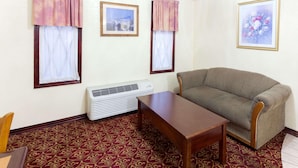Quarto com sofá, mesa, carpete estampado, duas janelas com cortinas e quadros nas paredes.