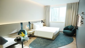 Quarto de hotel com cama, mesa de cabeceira, luminária, cadeira e vista para a cidade pela janela.