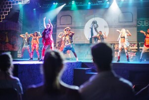 Um palco com artistas em diferentes figurinos, um fundo de parede de tijolos e uma grande tela iluminada.