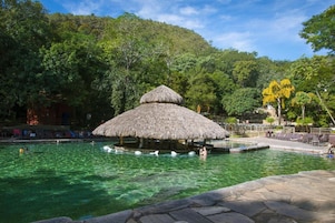 Uma piscina com estrutura de telhado de palha, cercada por vegetação exuberante e uma colina ao fundo.