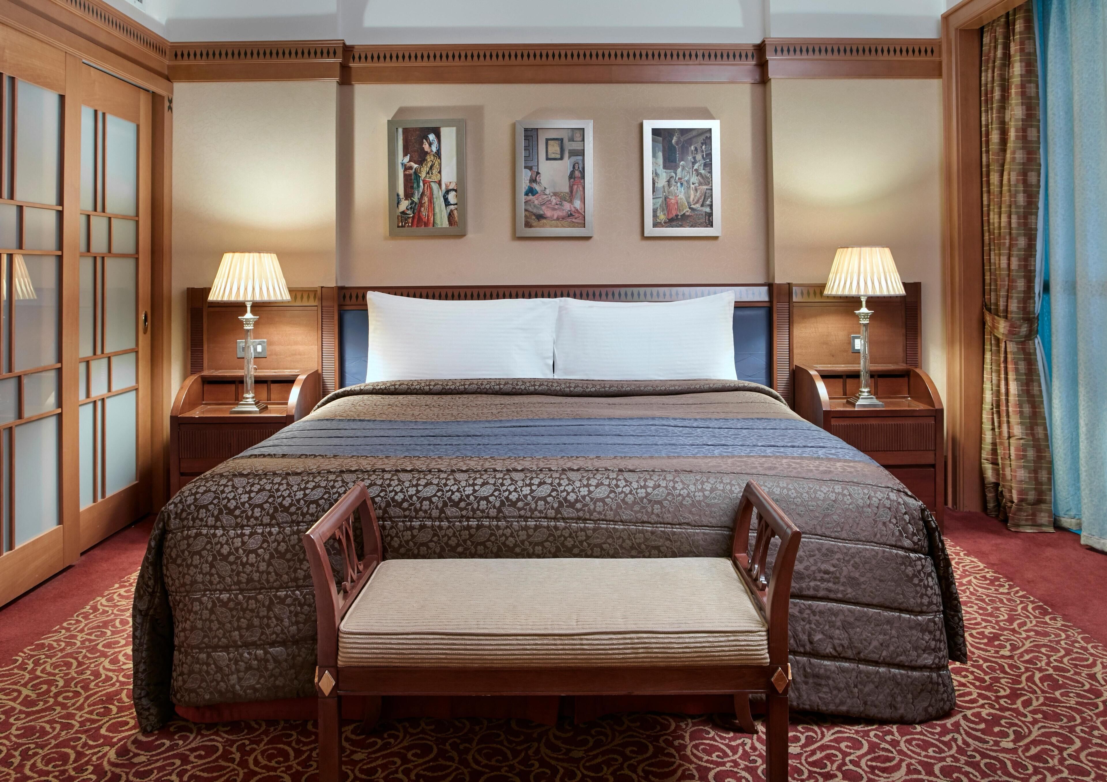 Quarto de hotel com uma cama grande, duas mesas de cabeceira com abajures, uma poltrona e três obras de arte emolduradas na parede.