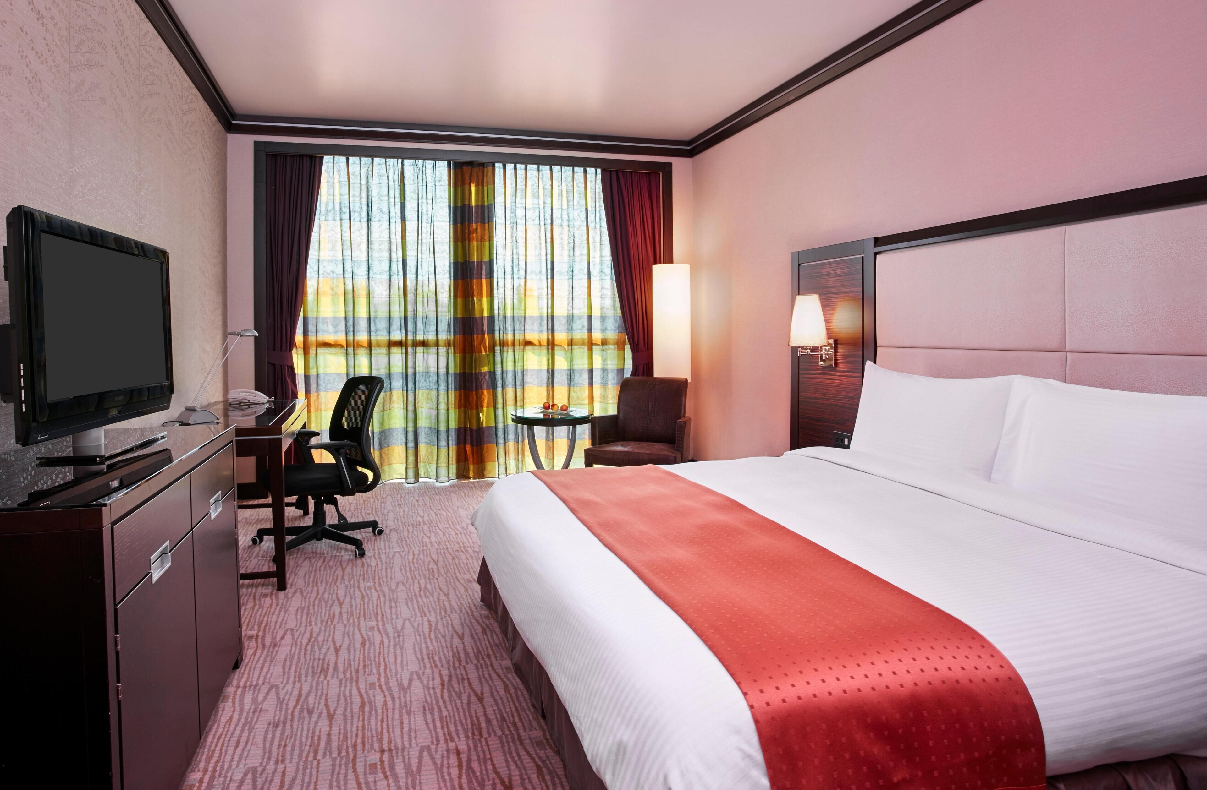 Quarto de hotel com cama, escrivaninha, cadeira, televisão e cortinas.