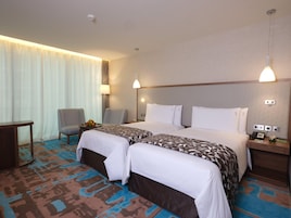 Quarto de hotel com duas camas, uma escrivaninha, uma cadeira, um abajur e uma cesta de frutas.
