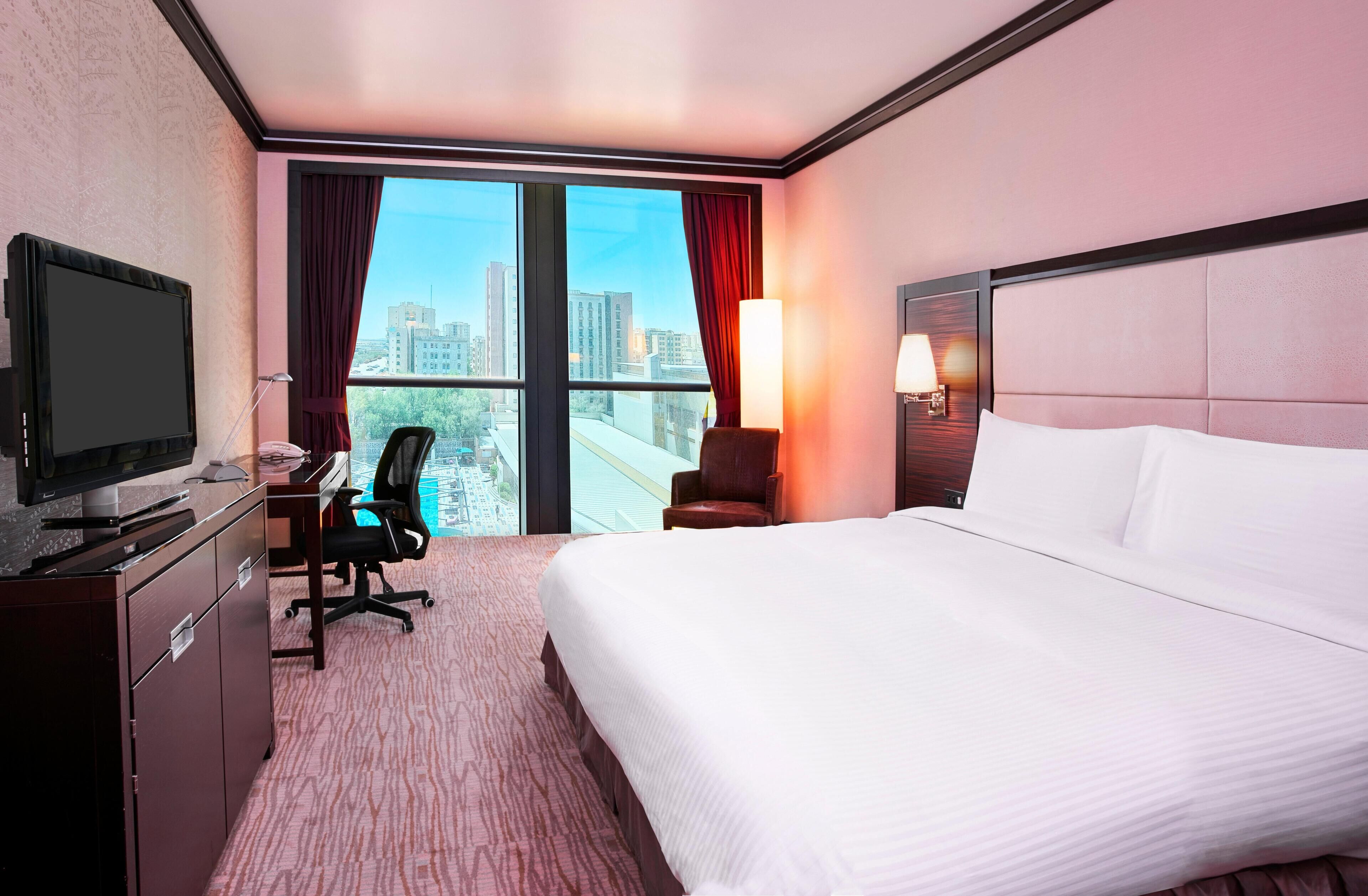 Quarto de hotel com cama, escrivaninha, cadeira, televisão e vista para a paisagem urbana.
