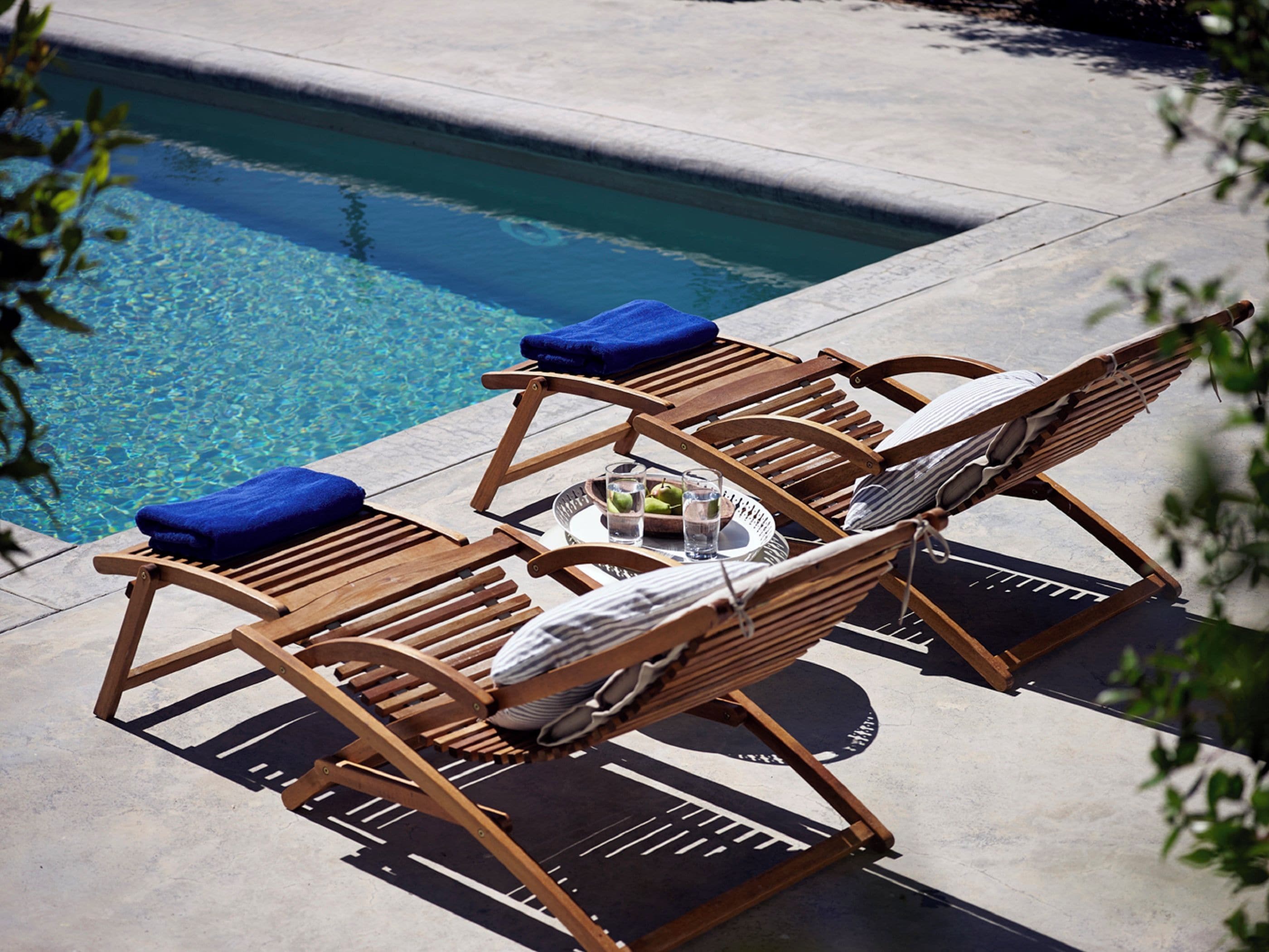 Uma área junto à piscina com espreguiçadeiras de madeira e uma mesa, com vista desobstruída para a piscina e piso em azulejo.